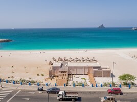 Um edifício à beira-mar com telhado plano, um caminho pavimentado que leva à praia e uma vista desobstruída do oceano e uma ilha distante.