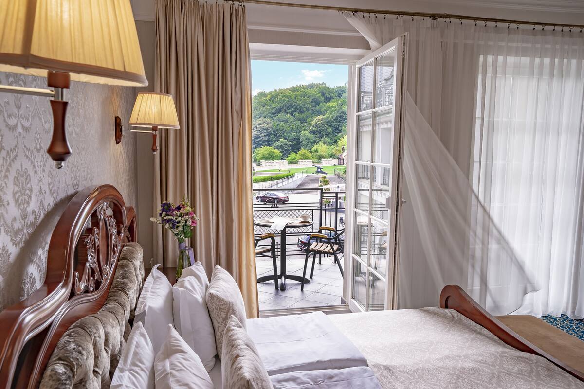 Quarto de hotel com cama, cabeceira de madeira, mesa de cabeceira, luminária e vista para uma varanda com mobiliário exterior.