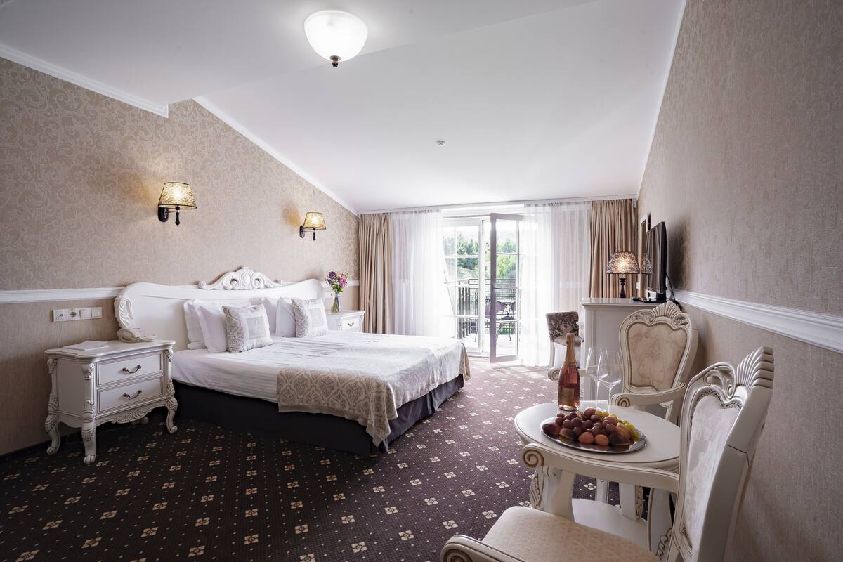 Quarto de hotel com uma cama grande, uma mesa de cabeceira, uma cadeira, uma mesa de jantar com um prato de frutas e uma varanda com cortinas.