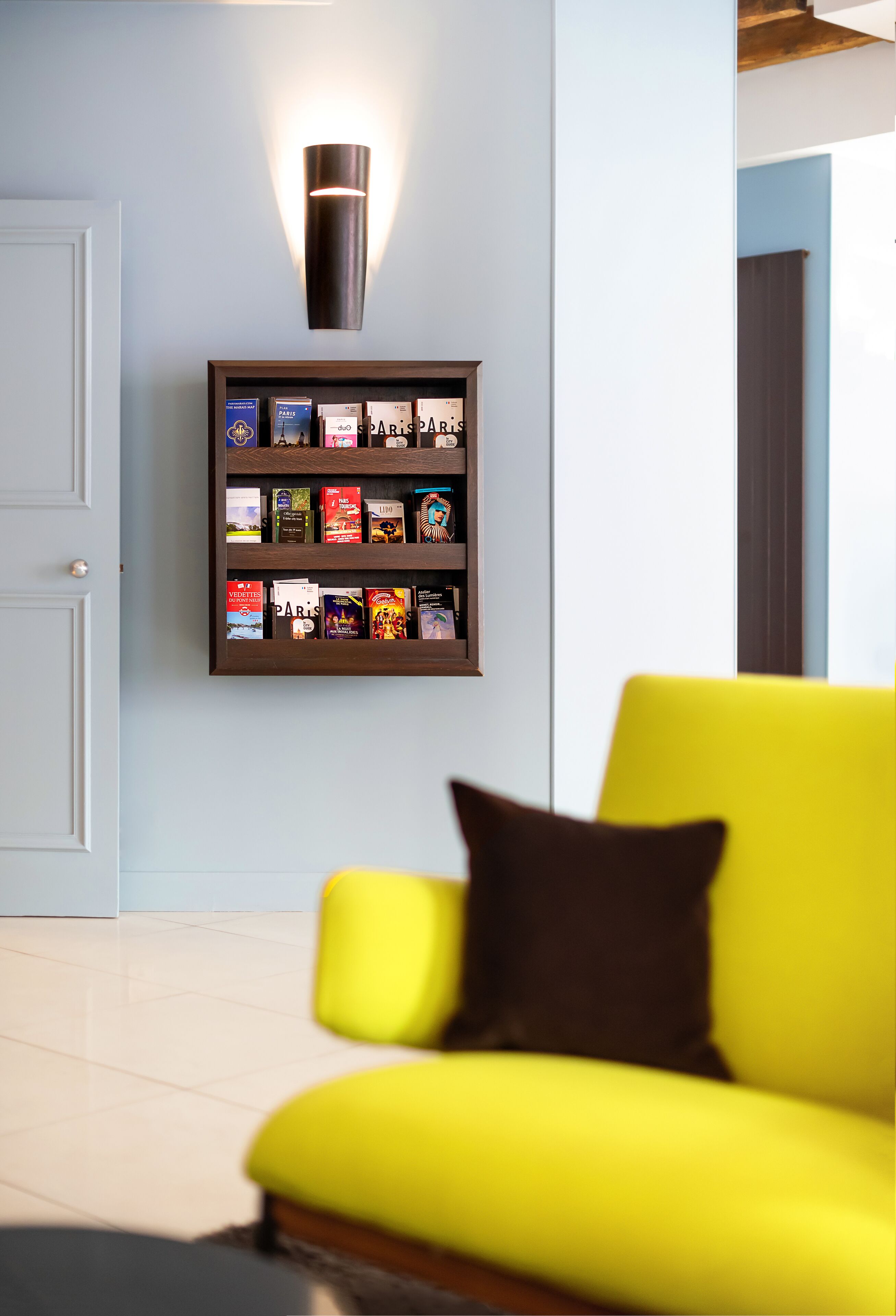 Uma sala de estar moderna com um sofá amarelo vibrante, uma estante fixada na parede com livros e uma porta ao fundo.