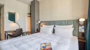 Quarto de hotel com uma cama grande, uma escrivaninha, uma cadeira, um espelho redondo e uma mesa de cabeceira com uma luminária.