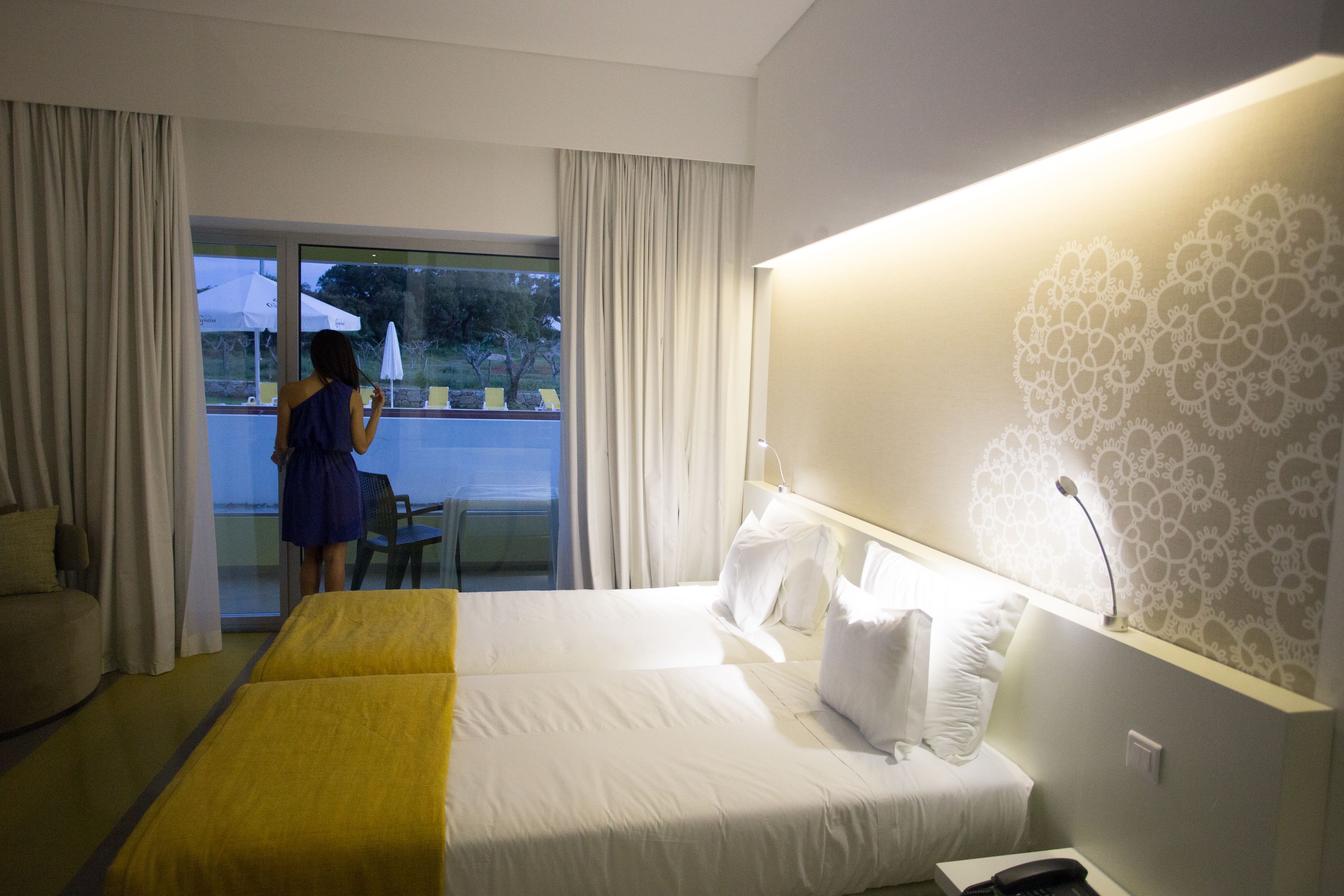 Quarto de hotel com cama, mesa de cabeceira, abajur e vista para o exterior.