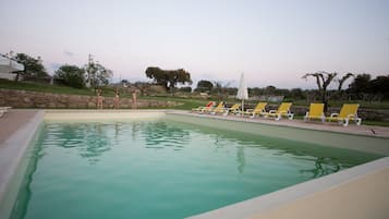 Piscina retangular com água cristalina, cercada por um muro de pedra e uma área gramada. Várias cadeiras de sol amarelas e guarda-sóis estão dispostos à beira da piscina.