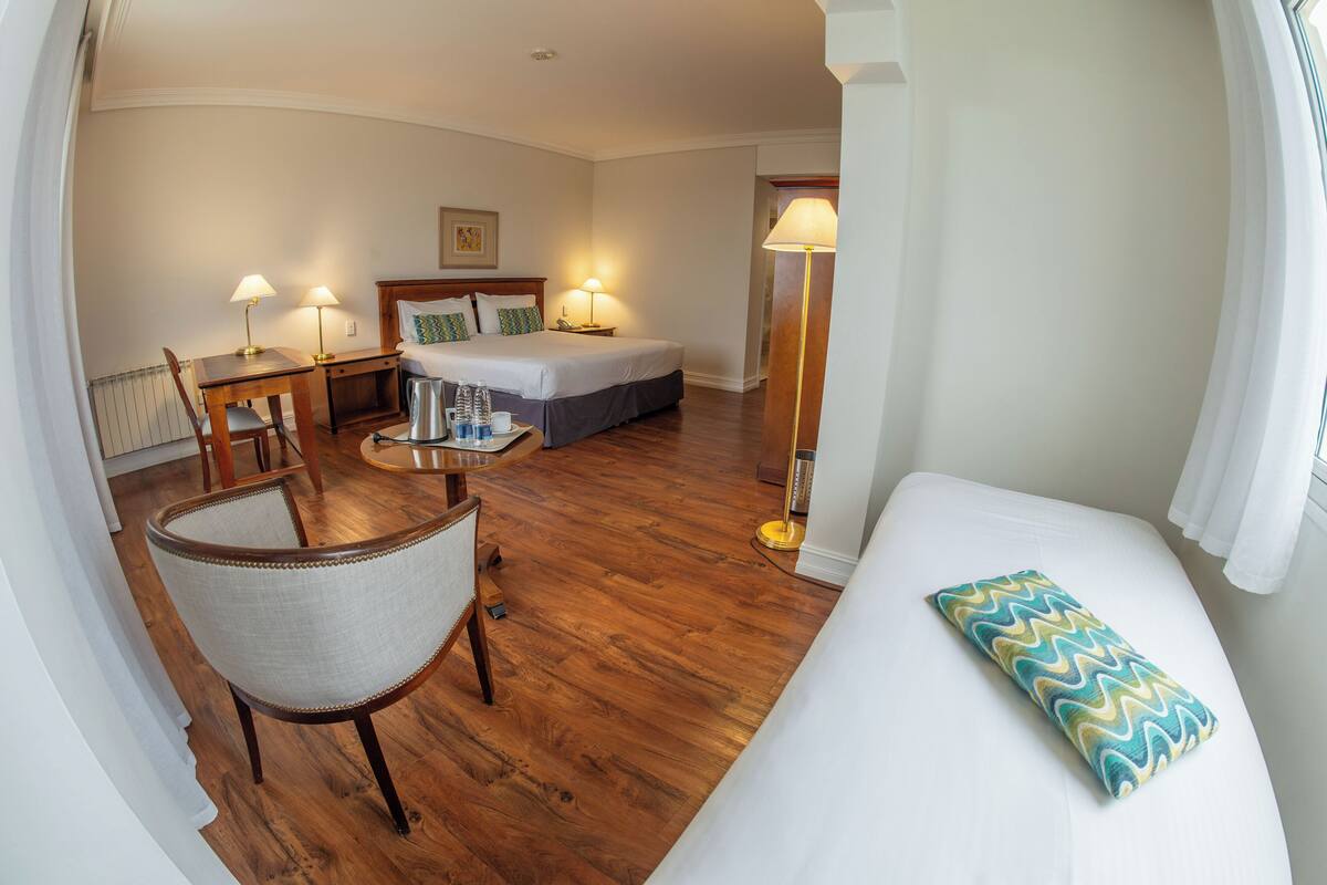 Quarto de hotel com cama, uma cadeira, escrivaninha e abajur.