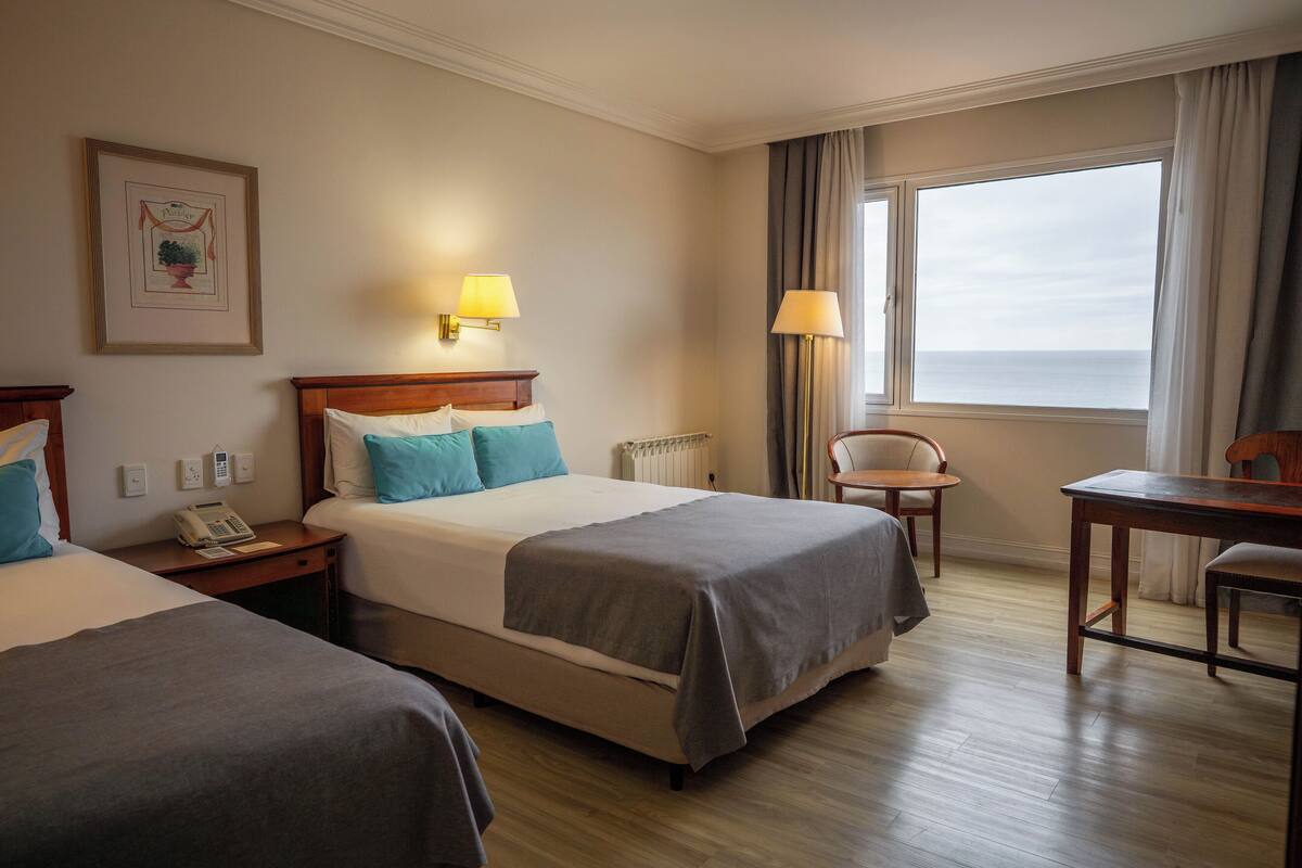Quarto de hotel com duas camas, uma escrivaninha e vista para o mar.