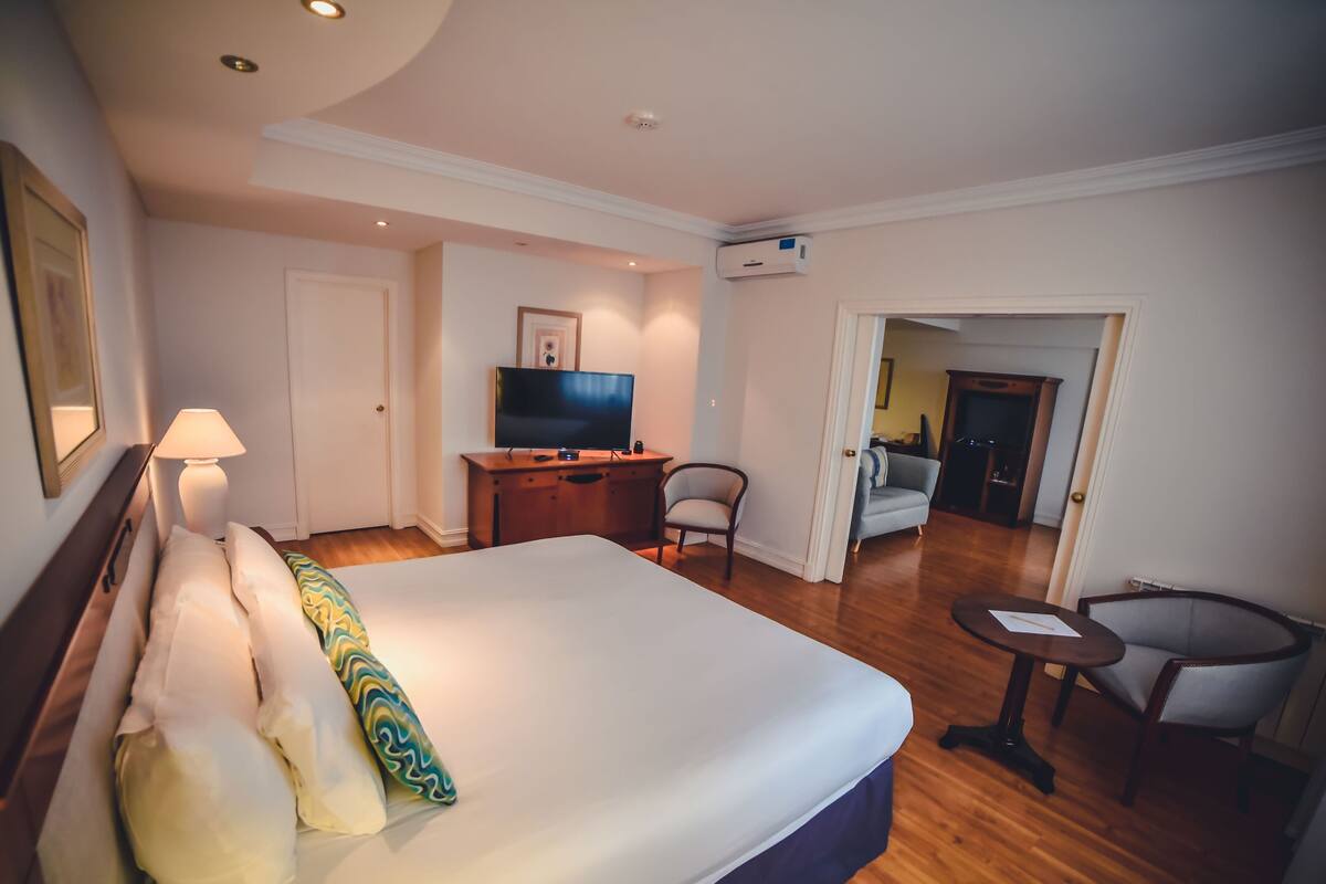 Quarto de hotel com uma cama grande, uma escrivaninha com televisão, uma cadeira e uma pequena mesa com um bloco de notas.