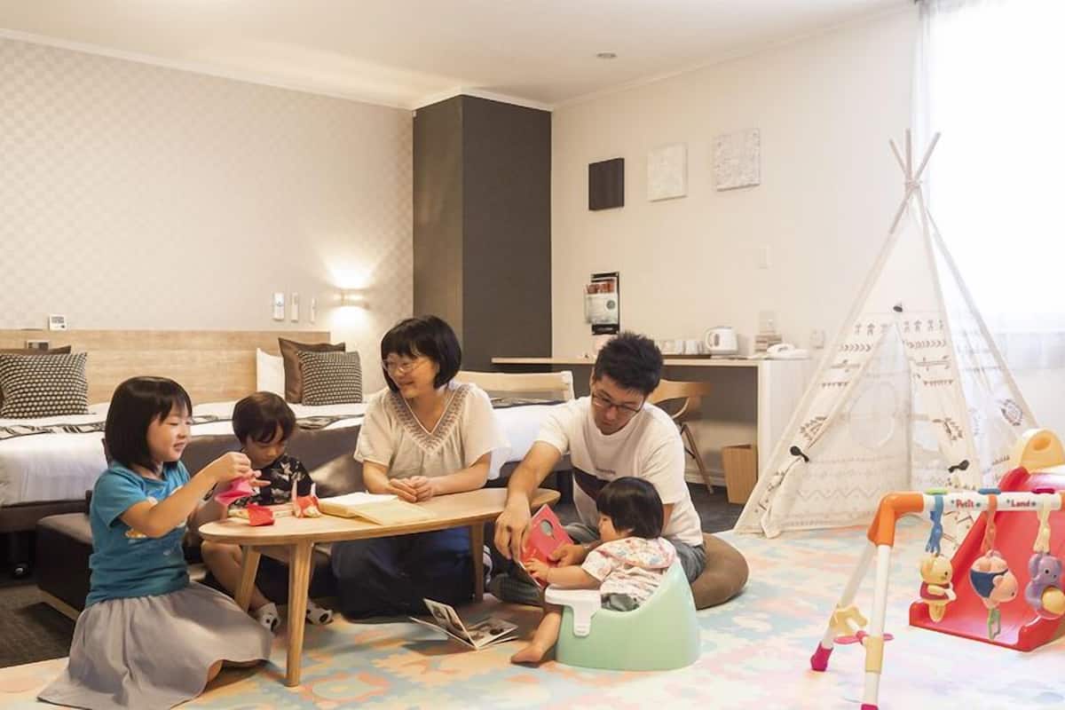 Uma família num quarto de hotel com uma cama, uma escrivaninha, uma cadeira, um espaço infantil com brinquedos e uma janela com cortinas.