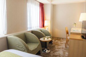 Quarto de hotel com duas poltronas verdes, uma mesinha redonda com luminária, escrivaninha de madeira e uma cama.