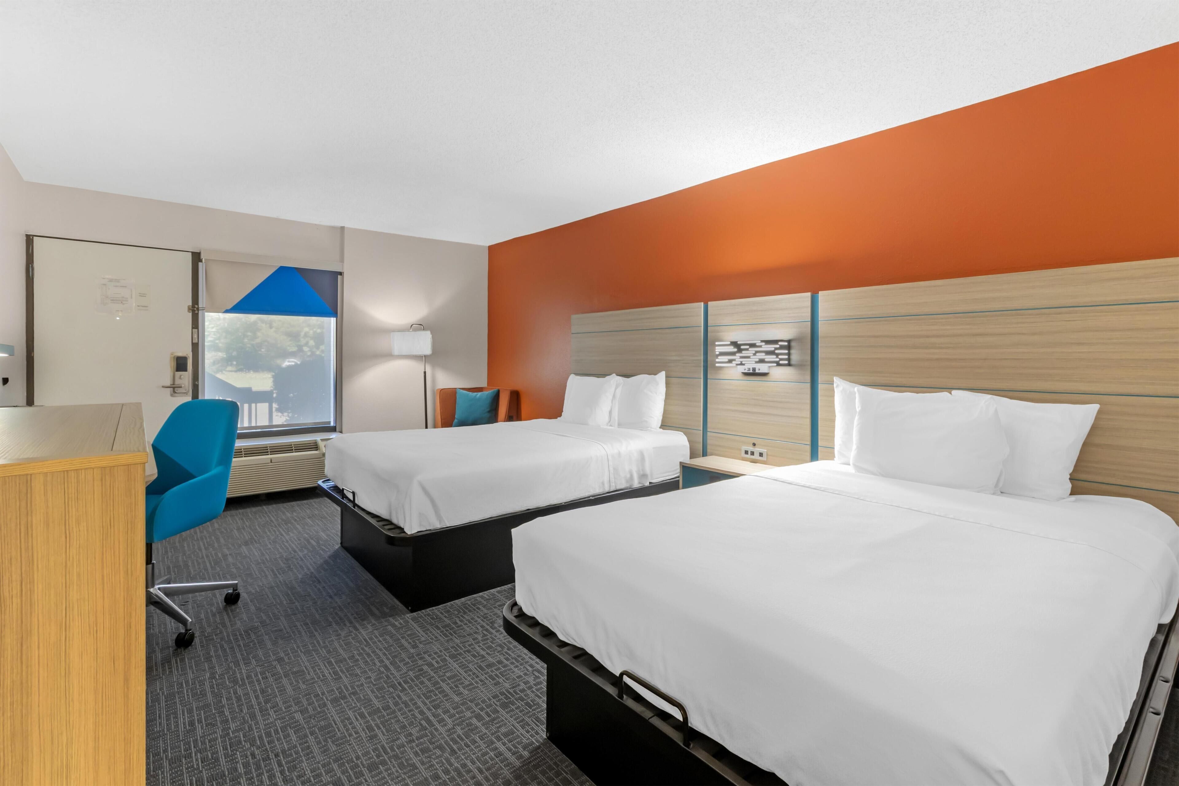 Quarto de hotel com duas camas, uma escrivaninha, uma cadeira e uma janela com vista.