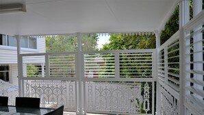 Área de varanda coberta com mesa de vidro e cadeiras de vime, delimitada por grades de metal branco ornamentadas.