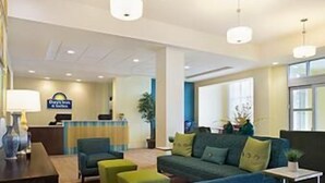 Um hall de hotel com balcão de recepção, área de estar com sofás e pufes, e uma grande janela que oferece iluminação natural.