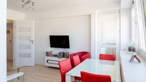 Uma sala de estar moderna com uma mesa de vidro e cadeiras vermelhas, uma televisão de tela plana presa na parede e uma porta branca que dá para outro cômodo.