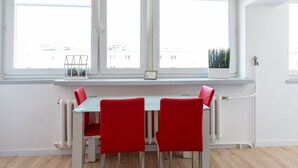 Uma área de refeições com uma mesa e quatro cadeiras vermelhas, uma janela com vista para prédios e um vaso de planta no parapeito.