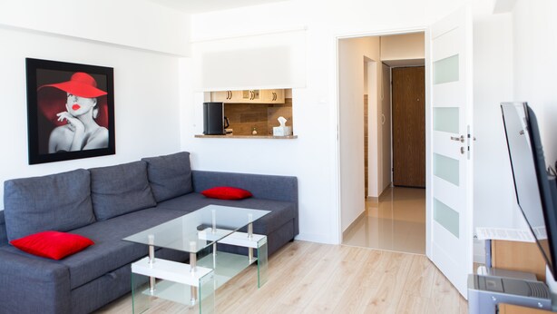 Uma sala de estar moderna com um sofá cinza, almofadas vermelhas, uma mesa de centro de vidro e uma área de cozinha com armários de madeira e bancada branca.
