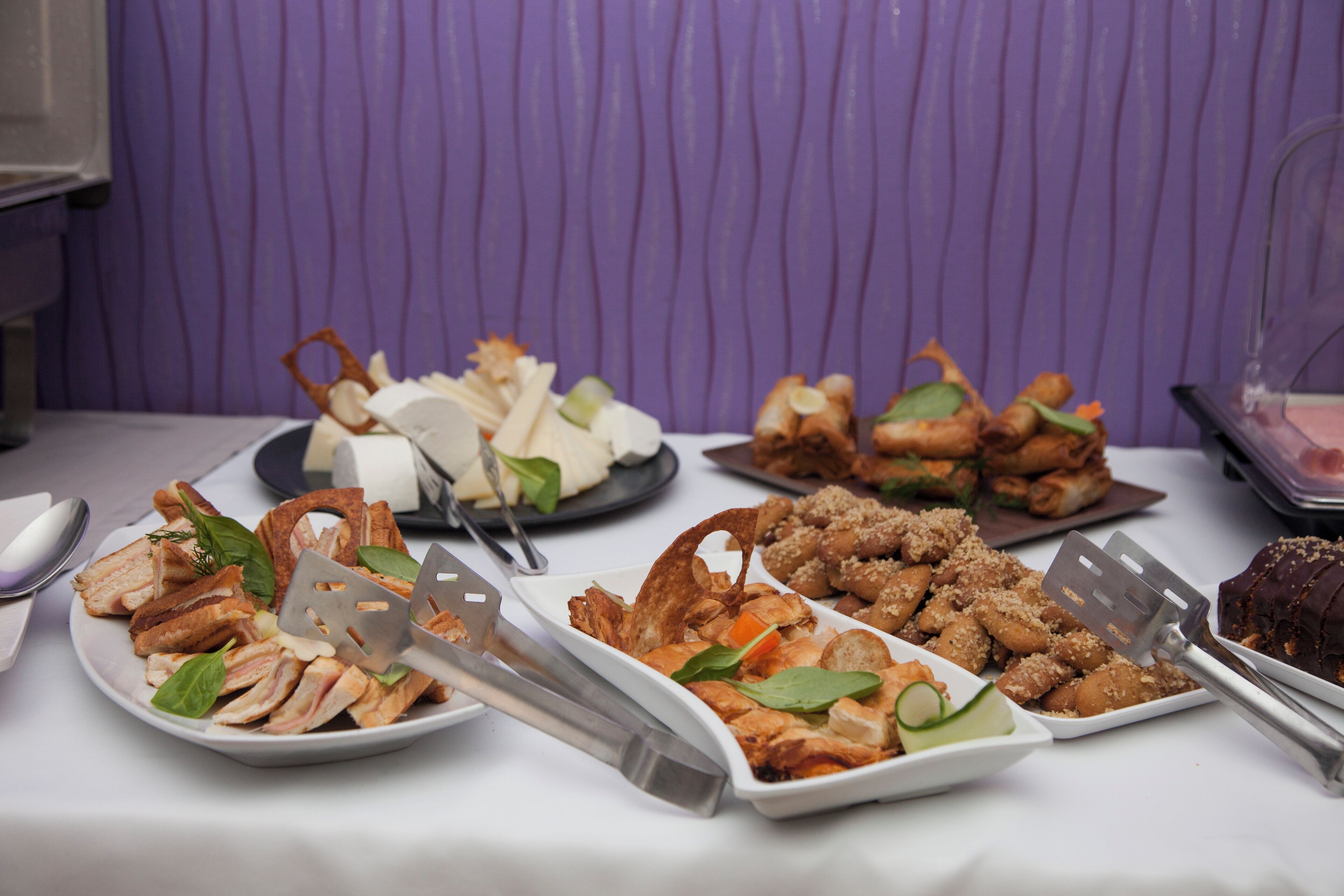 Uma mesa de buffet com diversos pratos, incluindo sanduíches, saladas e sobremesas, e utensílios de servir.