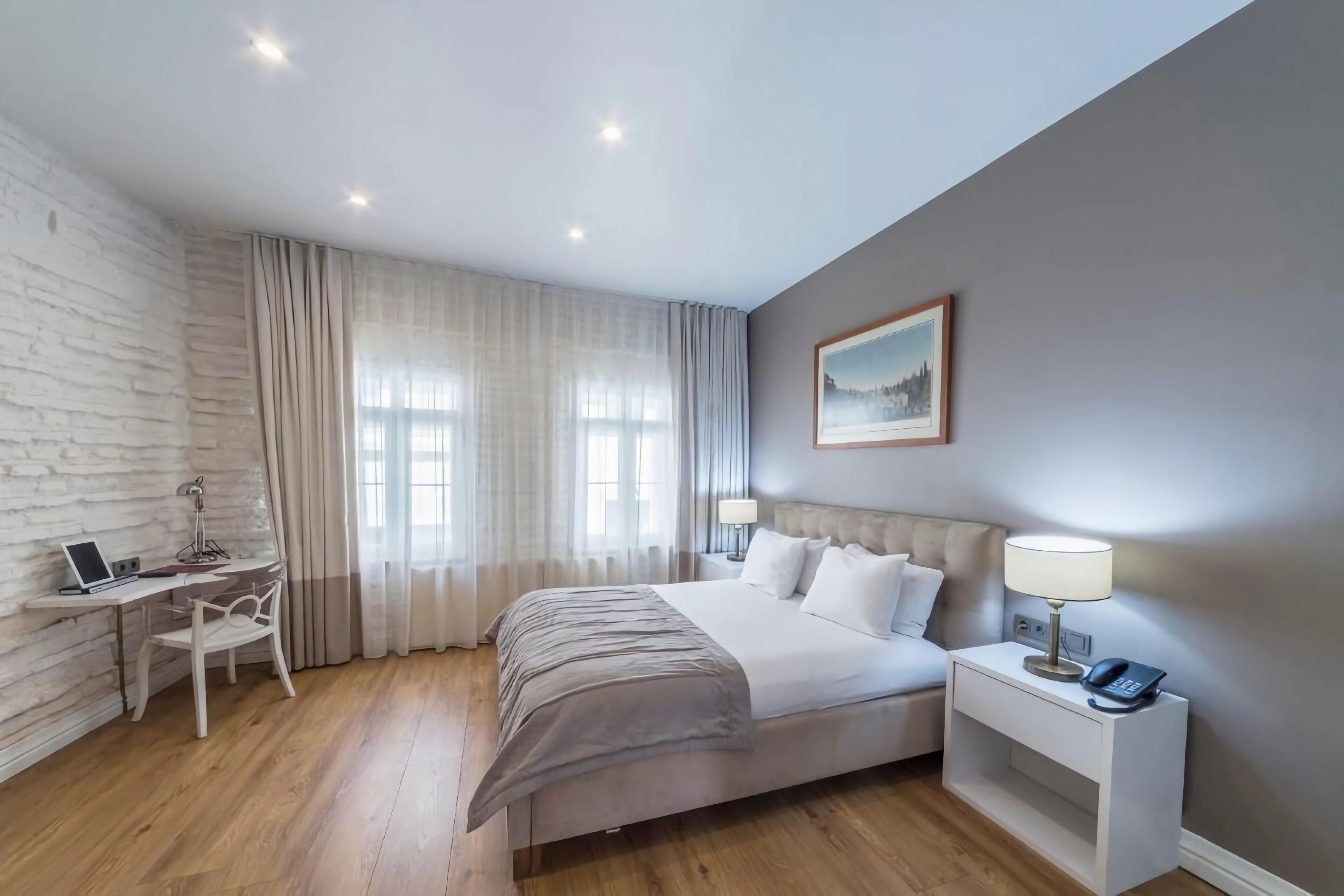 Quarto de hotel com cama, escrivaninha com laptop, cadeira, mesa de cabeceira com abajur e um quadro na parede.