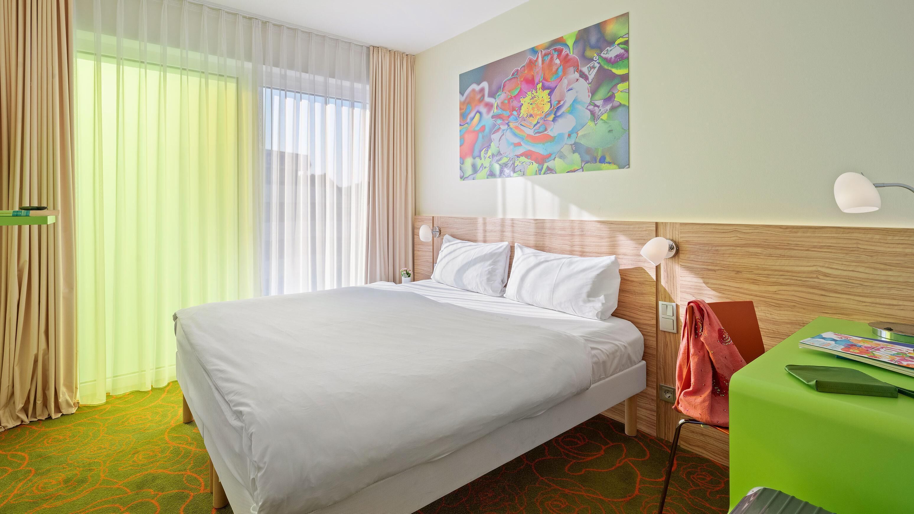 Quarto de hotel com uma cama grande, um quadro na parede, uma escrivaninha com abajur e uma cadeira verde.