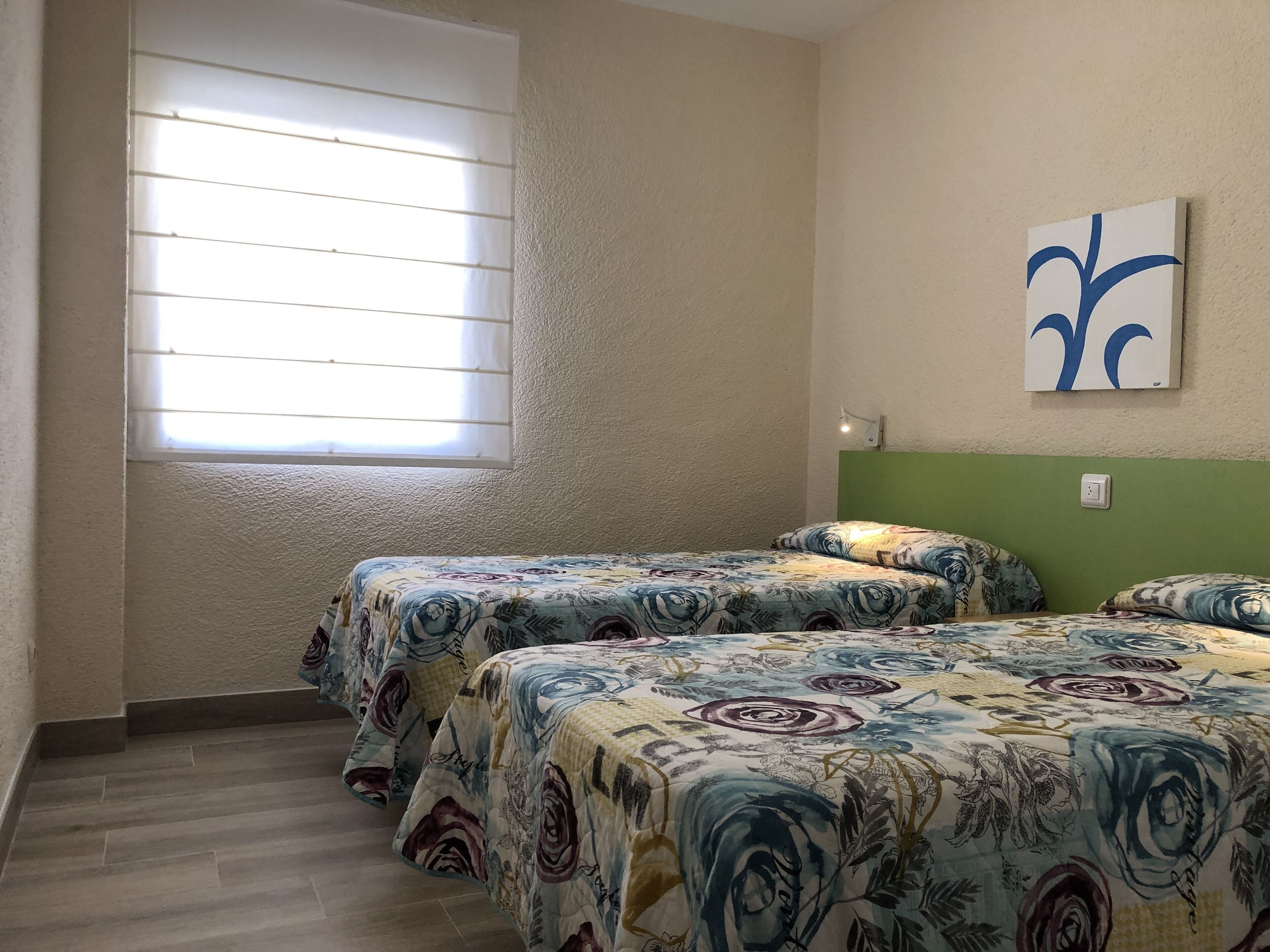 Quarto com duas camas, uma janela com persianas, cabeceira verde e luminária fixada na parede.