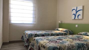 Quarto com duas camas, uma janela com persianas, cabeceira verde e luminária fixada na parede.