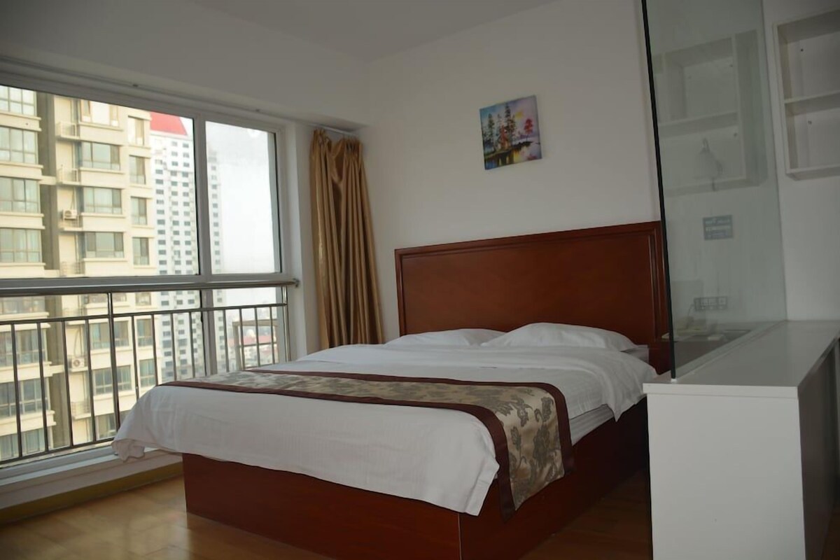 Quarto de hotel com cama, janela com vista para prédios, um quadro na parede e uma escrivaninha.