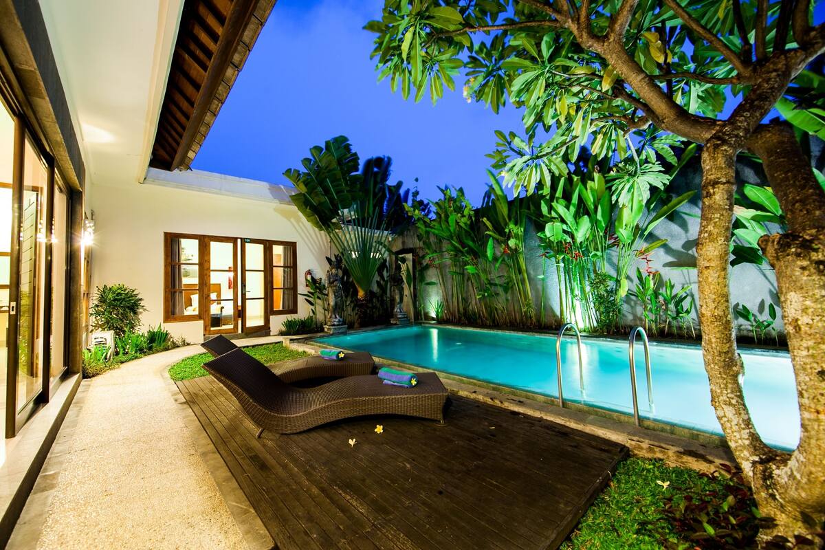 Uma área de piscina ao ar livre moderna, com deck de madeira, espreguiçadeiras e plantas tropicais.