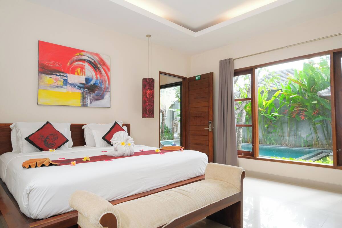 Quarto de hotel com uma cama grande, um banco, uma janela com vista para o verde e um quadro na parede.