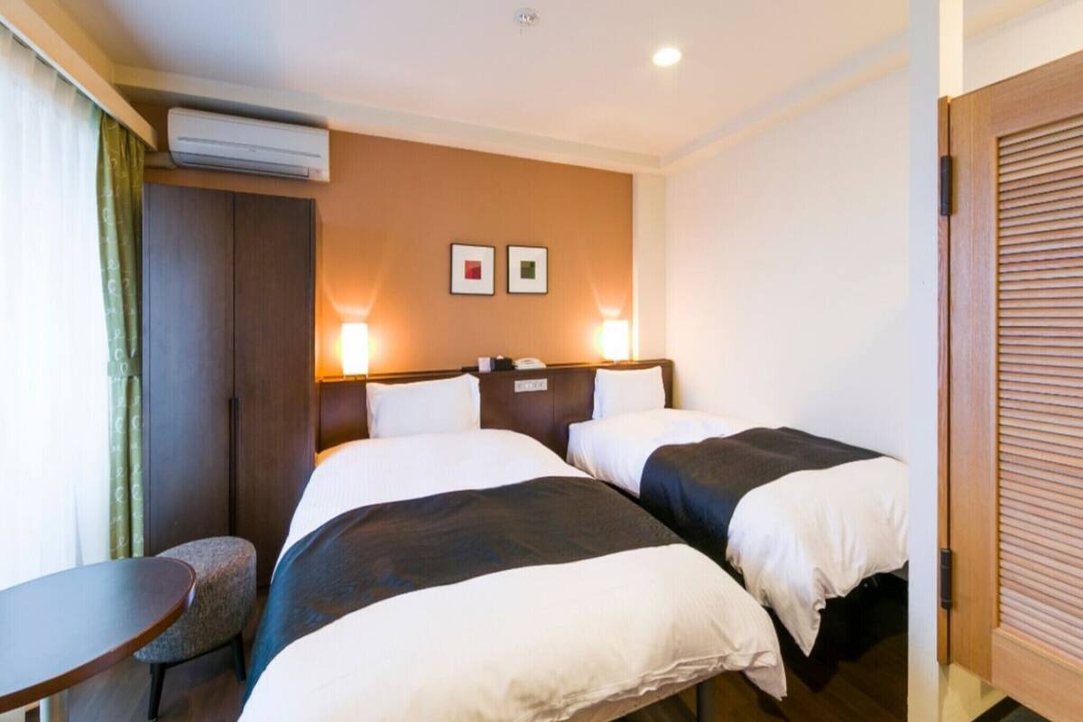 Quarto de hotel com duas camas, uma mesinha, uma cadeira e um ar condicionado.