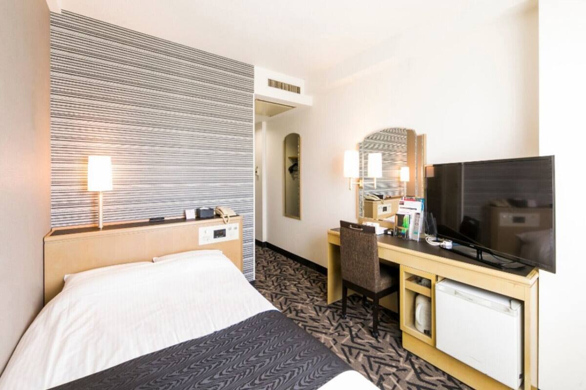Quarto de hotel com cama, escrivaninha com televisão, cadeira e pequeno armário.
