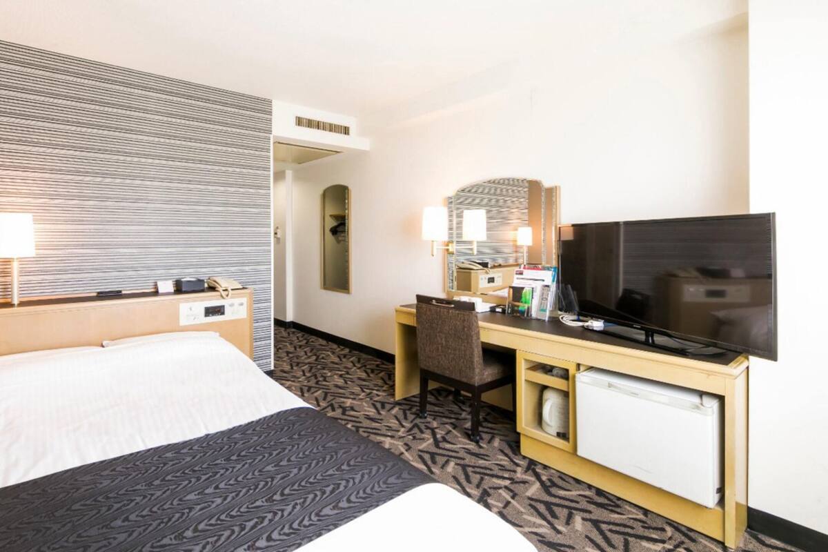 Quarto de hotel com cama, escrivaninha com televisão, uma cadeira e um espelho.