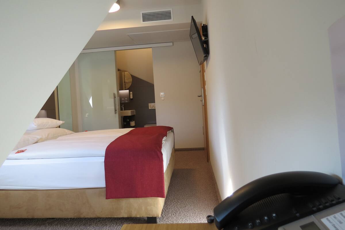 Quarto de hotel com cama, telefone e banheiro visível através de uma porta aberta.