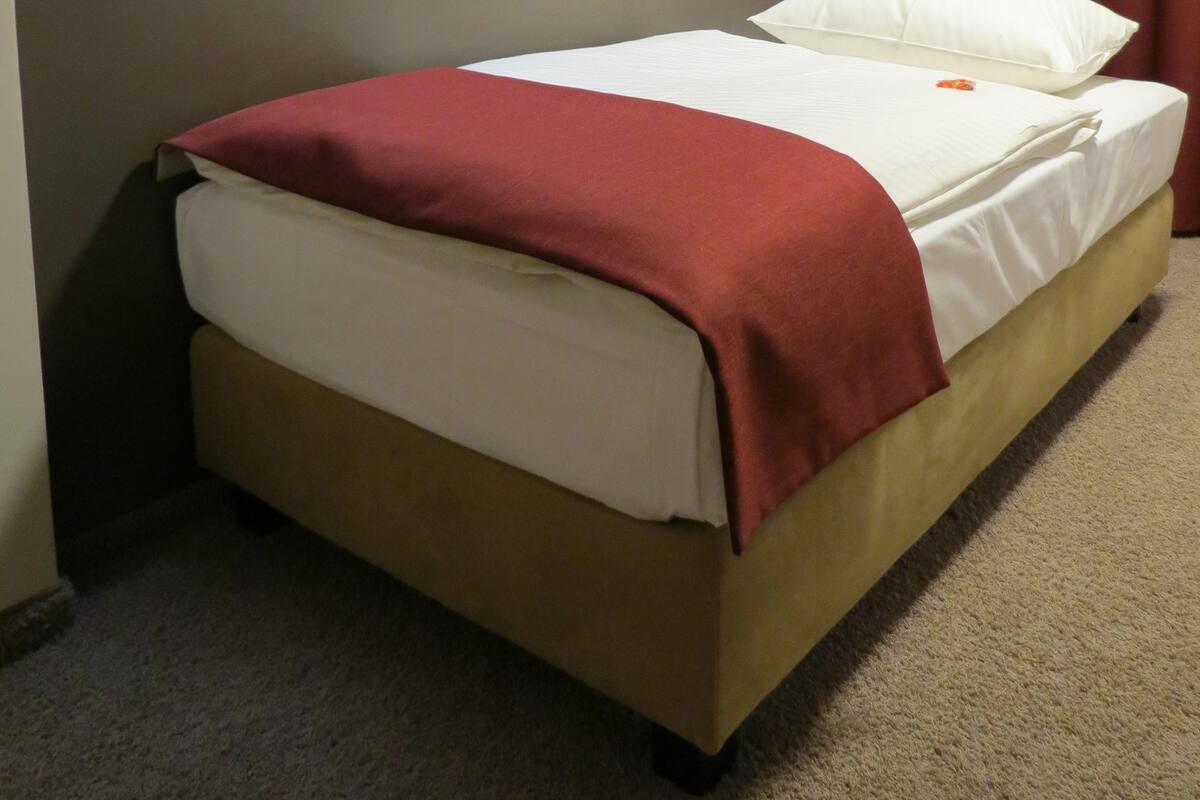 Uma cama de solteiro com um cobertor vermelho, um travesseiro branco e uma cabeceira bege em um quarto com carpete e cortina vermelha.