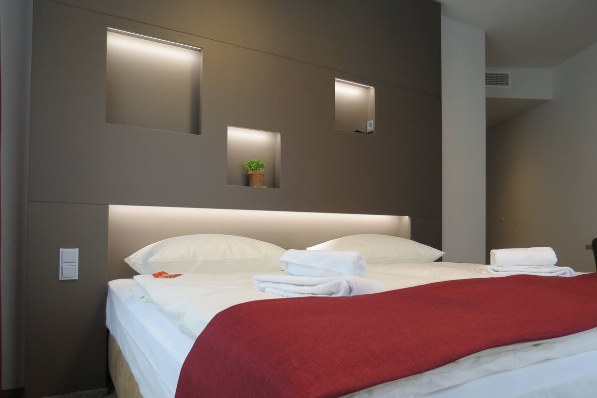 Quarto de hotel com cama grande, duas mesas de cabeceira, manta vermelha e iluminação na parede.