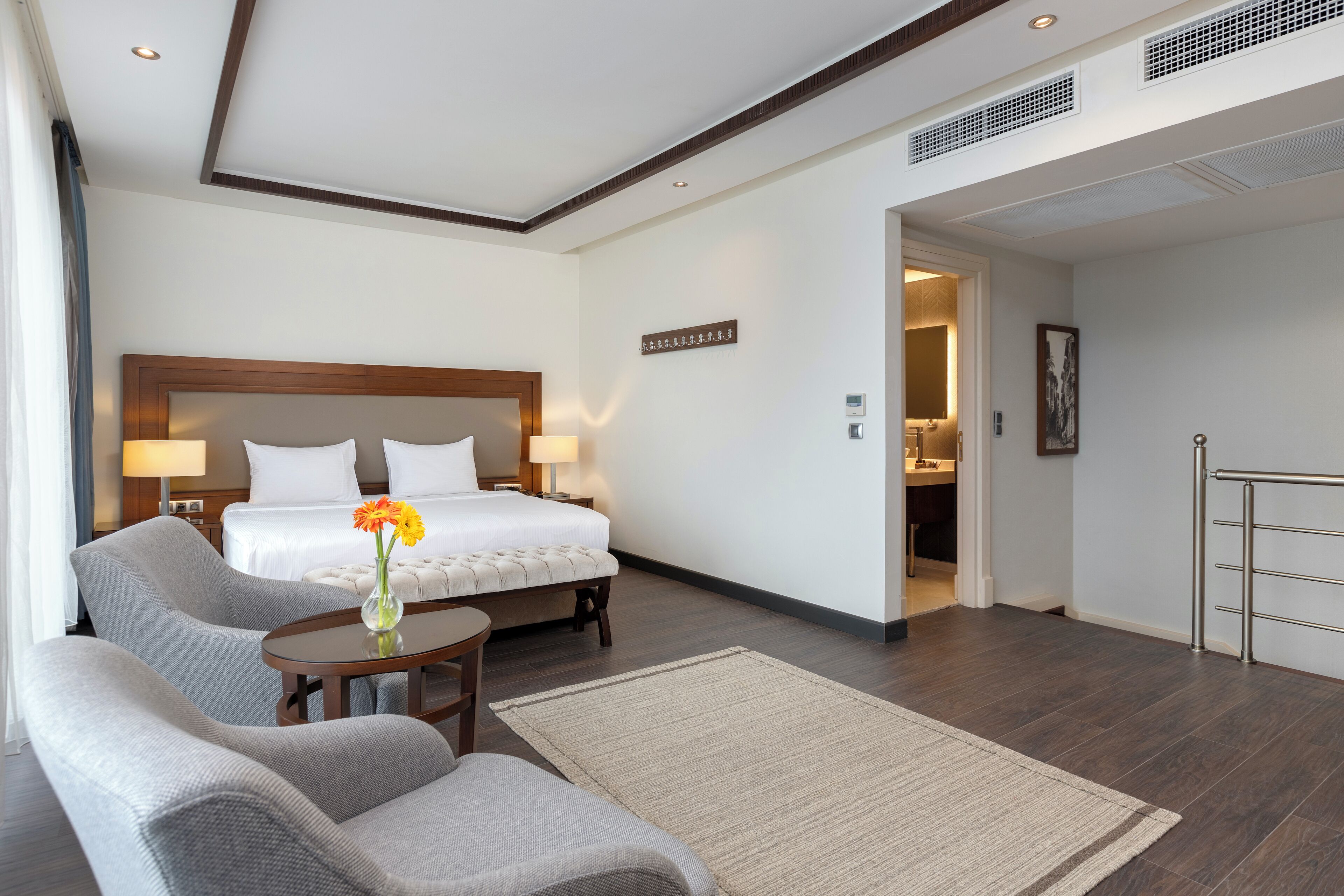Quarto de hotel com uma cama grande, duas mesas de cabeceira com luminárias, um espaço de estar com uma cadeira e uma mesinha, e vista para o banheiro.