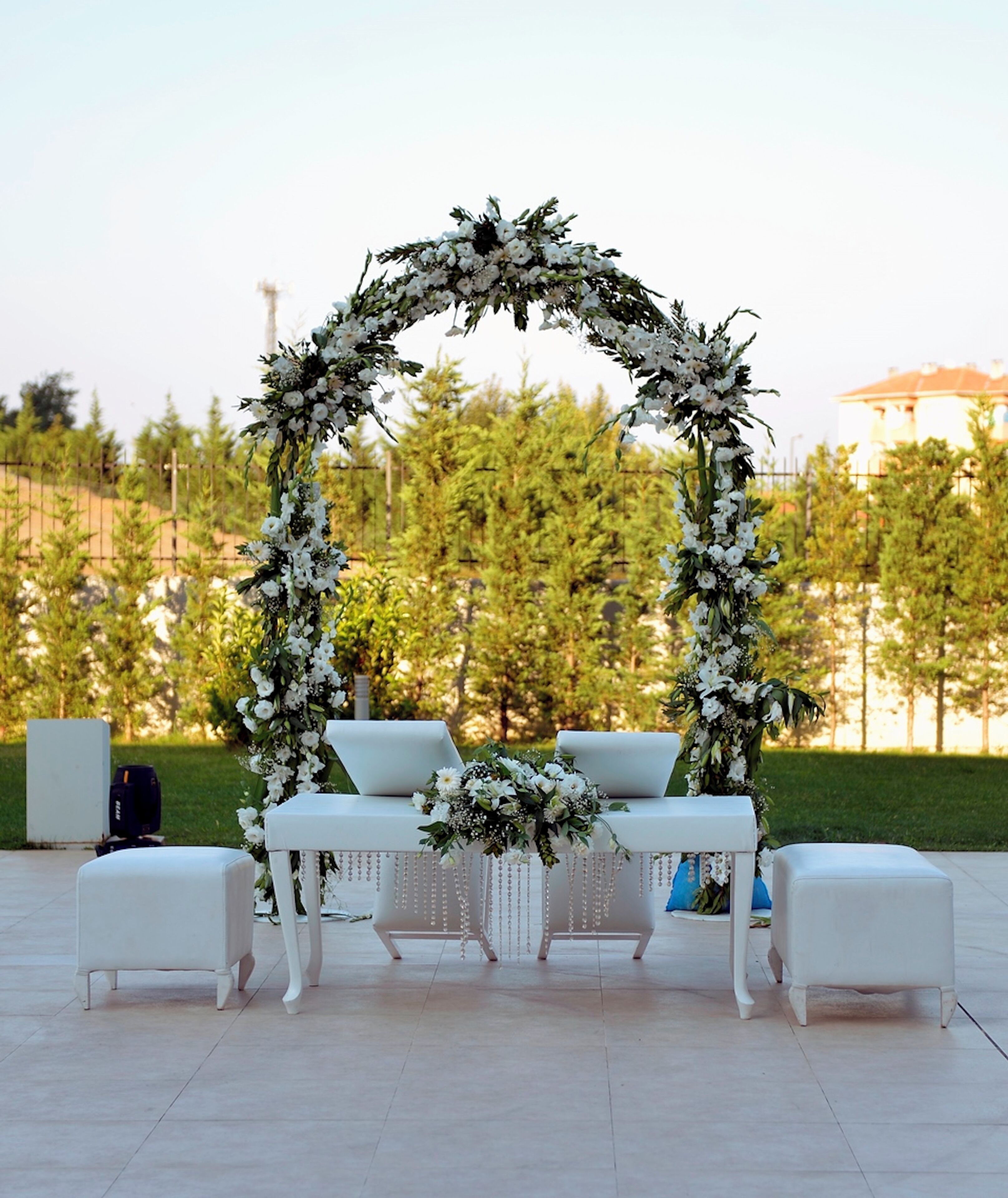 Decoração de casamento ao ar livre com arco floral, móveis brancos e piso de azulejos.