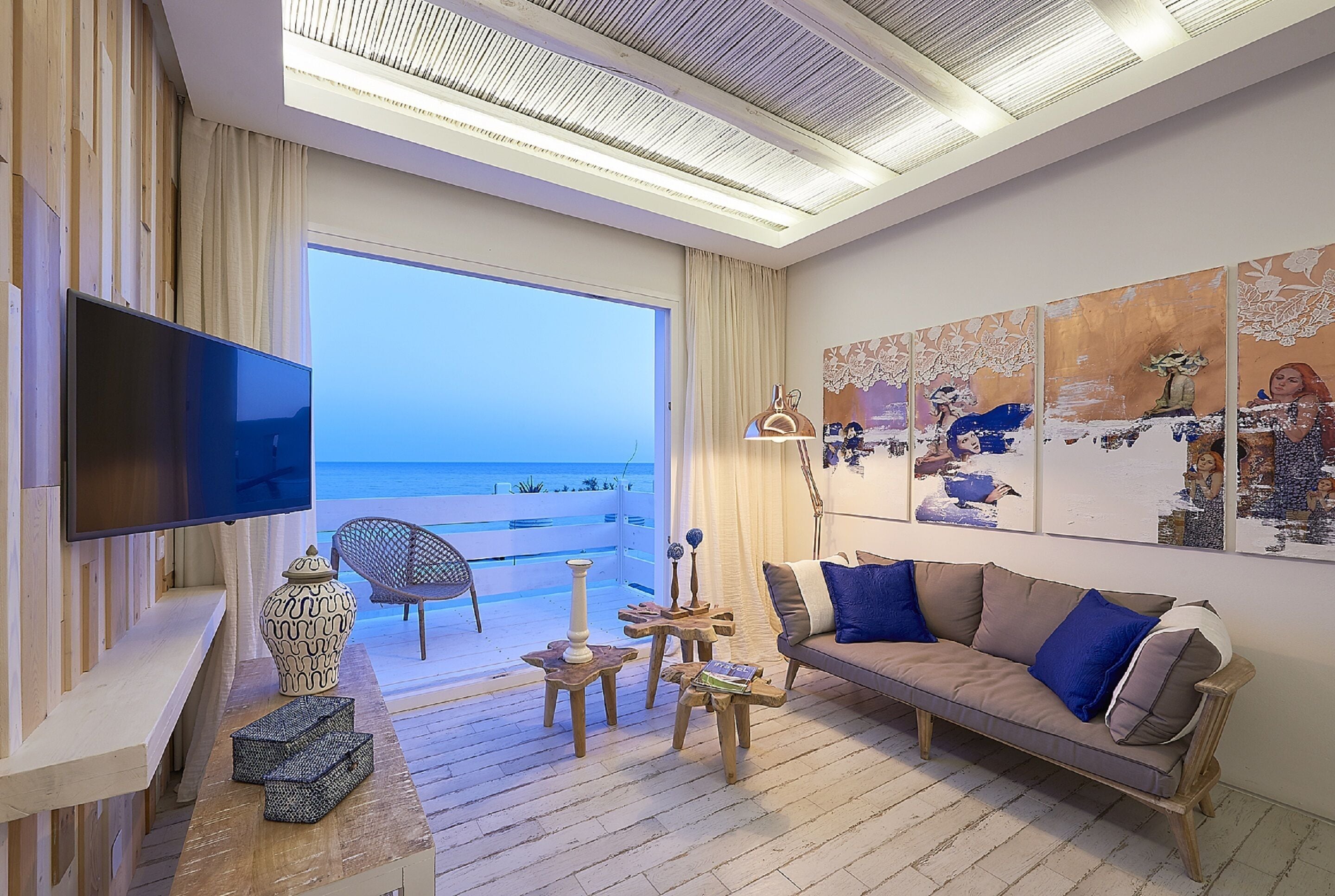 Uma sala de estar moderna com uma televisão grande, móveis de madeira, um sofá com almofadas azuis e vista para o mar.
