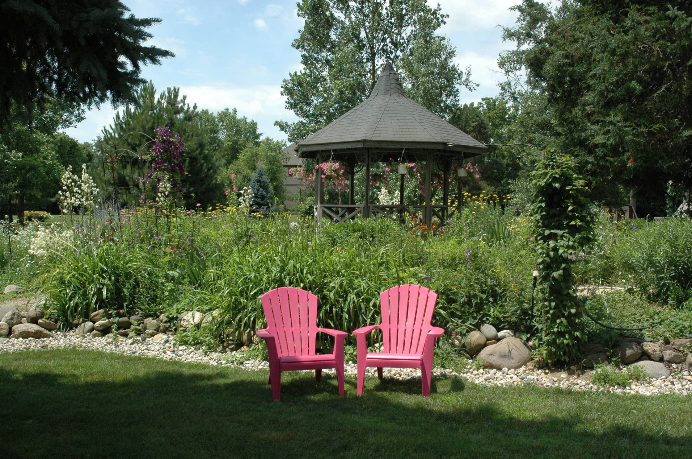 Um gazebo com telhado cónico rodeado por vegetação exuberante e duas cadeiras Adirondack à frente.