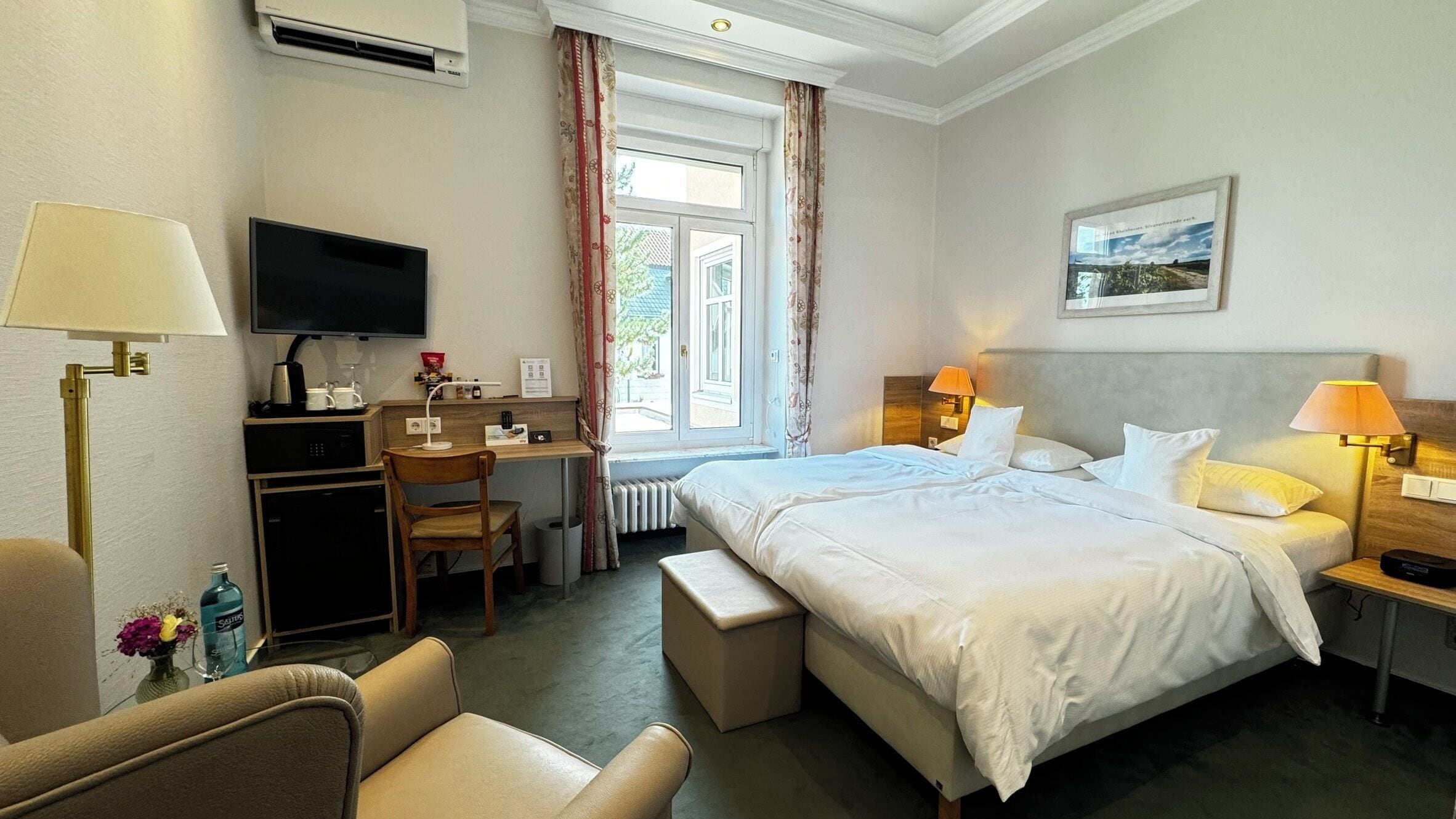 Quarto de hotel com cama, escrivaninha, cadeira, televisão e janela com cortinas.