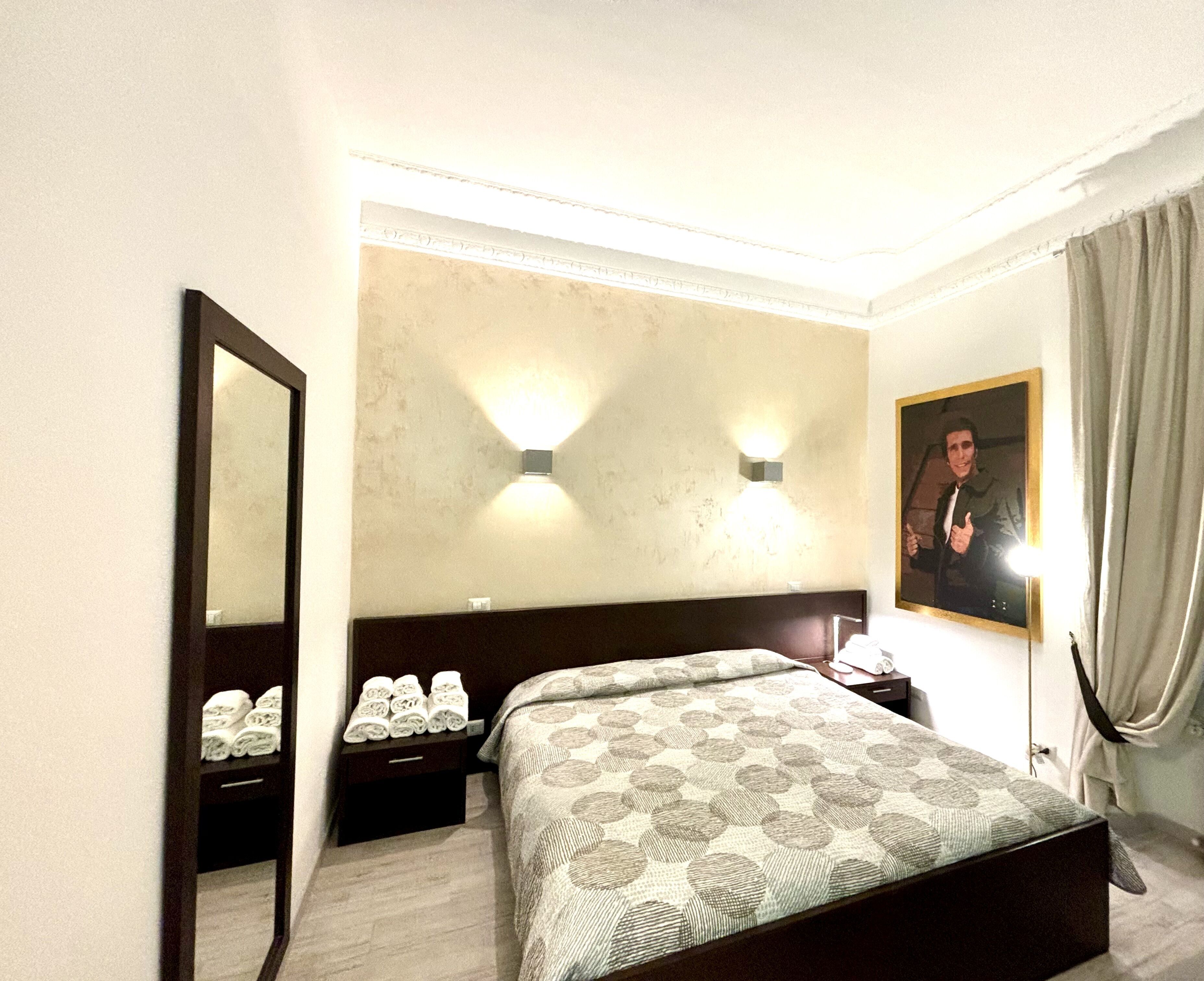 Quarto de hotel com uma cama grande, duas luminárias fixadas na parede, um espelho, uma mesa de cabeceira e um quadro emoldurado na parede.