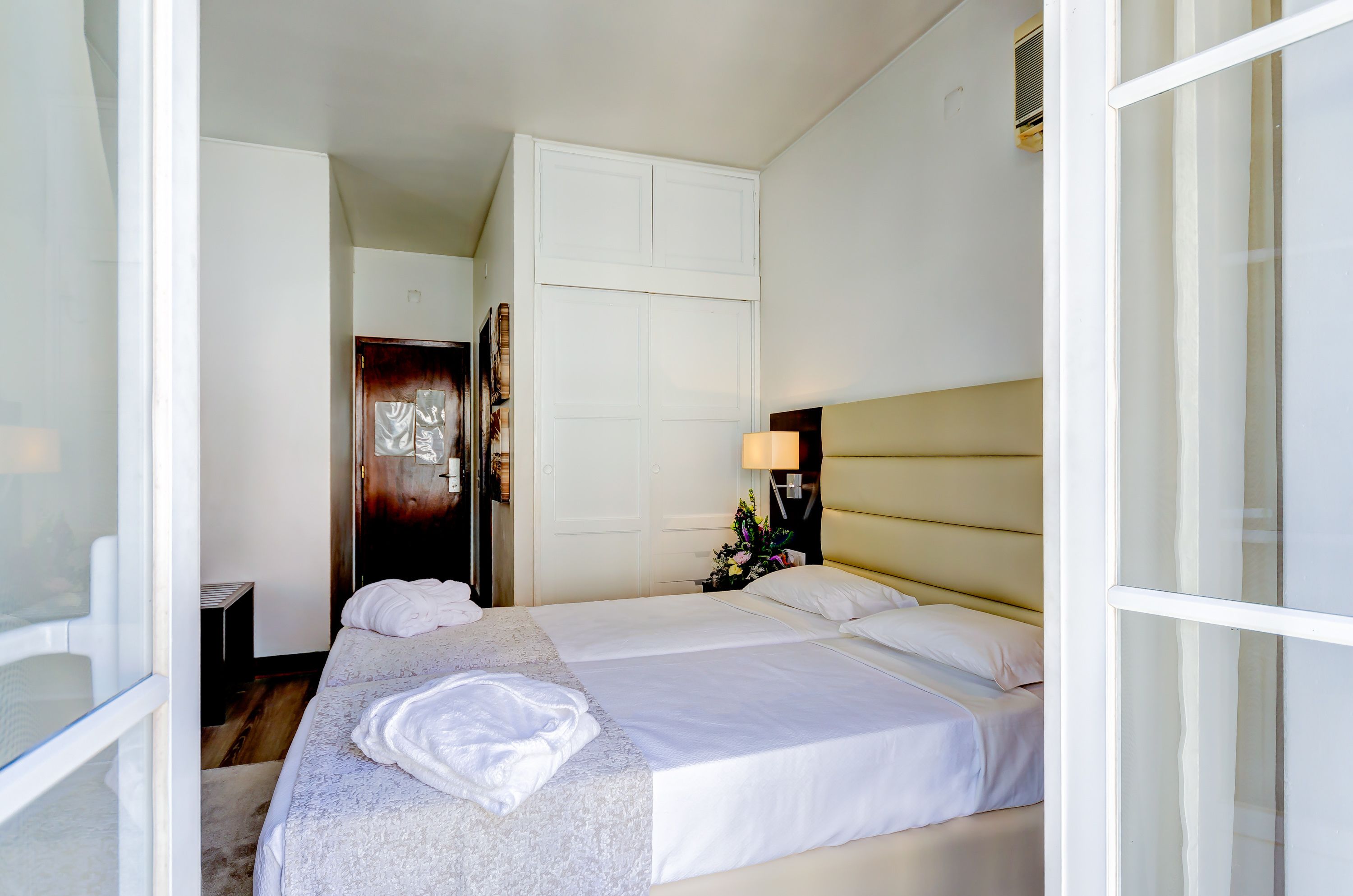 Quarto de hotel com cama, mesa de cabeceira, luminária e guarda-roupa.