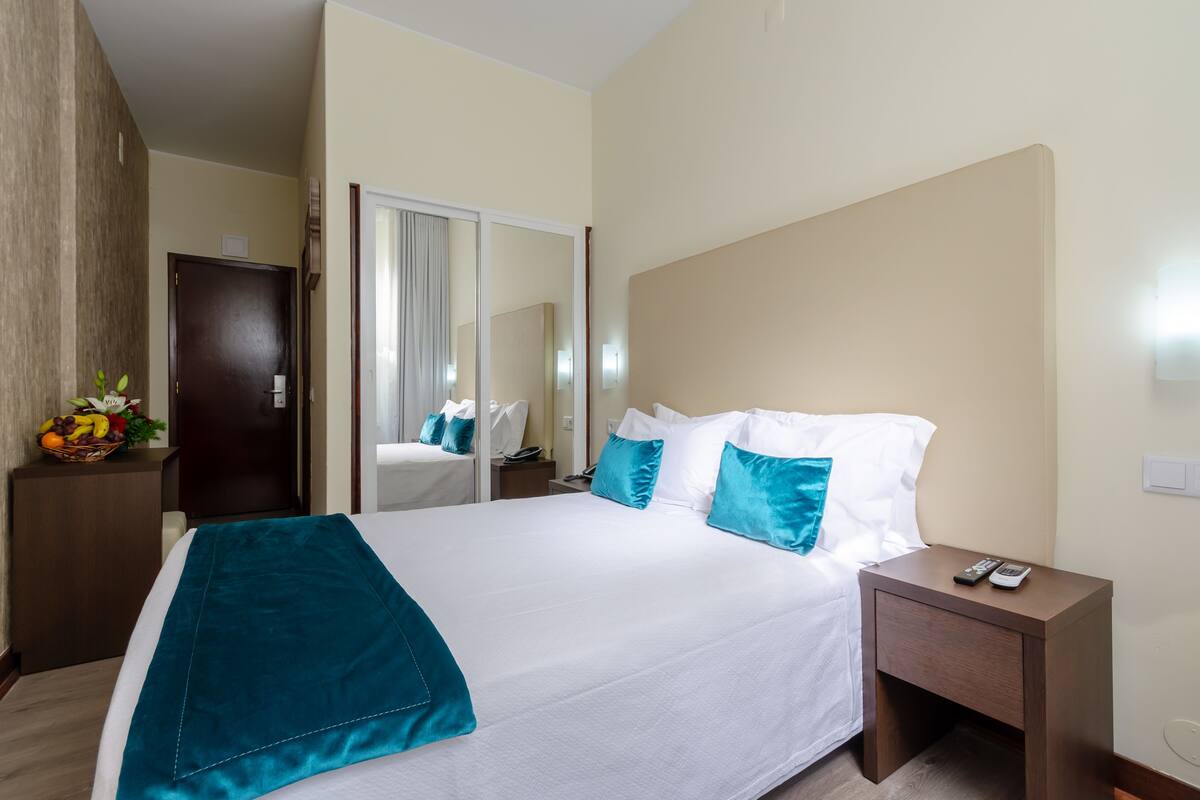 Quarto de hotel com uma cama grande, mesa de cabeceira com controle remoto, espelho e porta que dá para outro quarto.