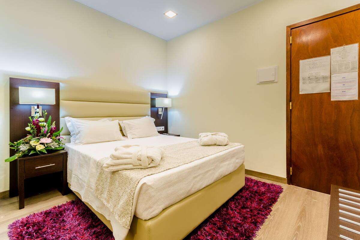 Quarto de hotel com cama, mesas de cabeceira, porta de madeira e um quadro de avisos.