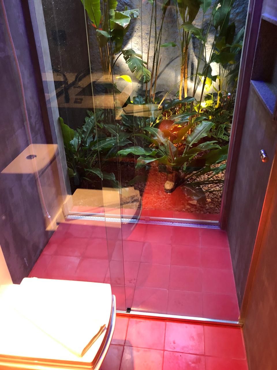 Quarto com chão de azulejos vermelhos, porta de vidro que dá para um jardim com plantas e um pequeno espelho d'água.