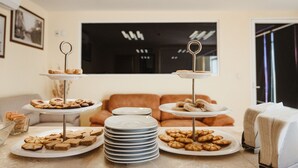 Disposição de buffet com várias travessas de doces e bolachas em suportes, uma pilha de pratos e um sofá ao fundo.