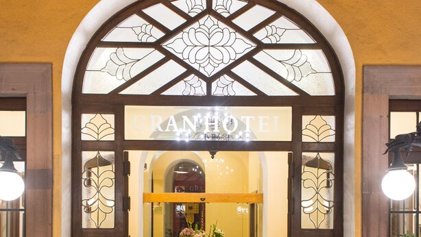 Entrada do Gran Hotel, com um tapete de boas-vindas, um arranjo floral e um padrão de piso xadrez.