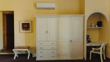 Quarto com um armário branco grande, um pequeno banco, uma escrivaninha com cadeira e um aparelho de ar condicionado na parede.