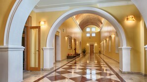 Um corredor de hotel com piso xadrez, teto em arco e uma série de portas ao longo das paredes.