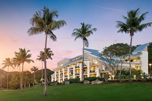 Um hotel moderno com vários andares, grandes janelas e um gramado bem cuidado com palmeiras.