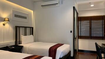 Quarto de hotel com duas camas, uma escrivaninha e uma cadeira. Há um ar condicionado na parede, um abajur no lado esquerdo e uma janela com persianas à direita.