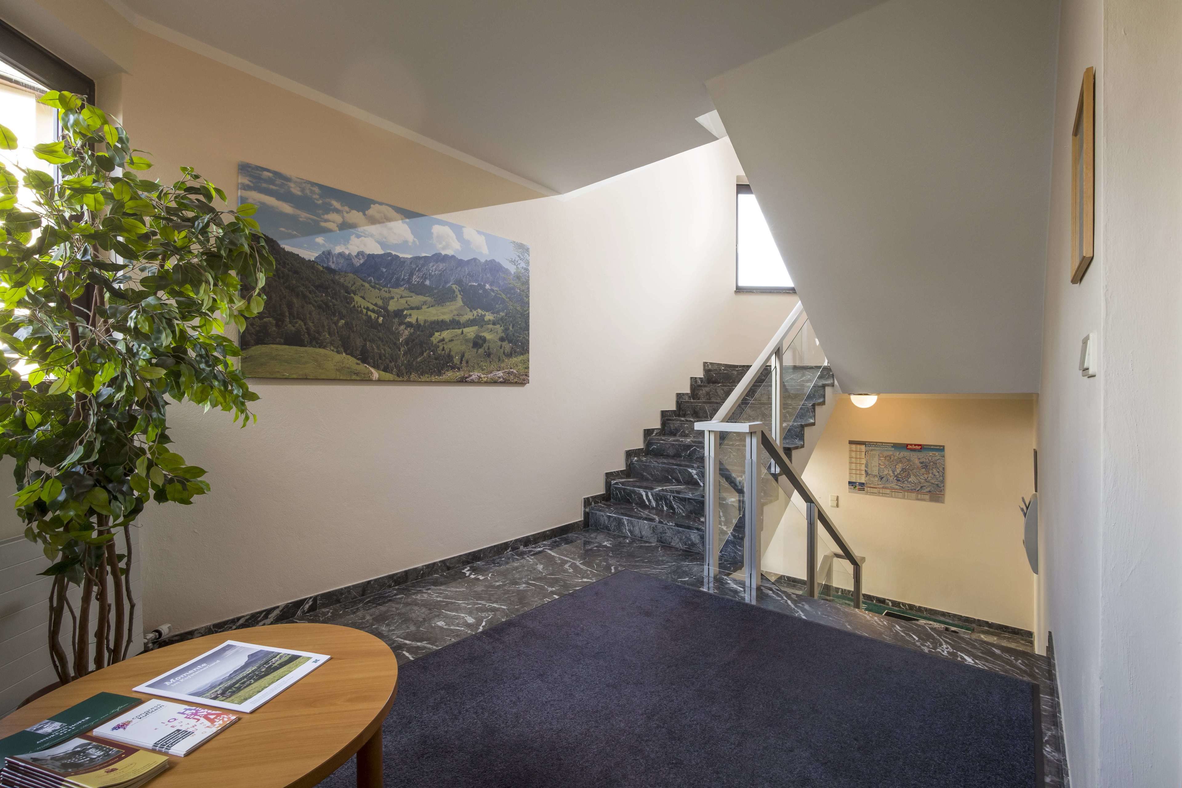Uma escadaria com degraus de mármore que leva a um andar superior, uma mesa de madeira com folhetos e um grande quadro com uma paisagem na parede.