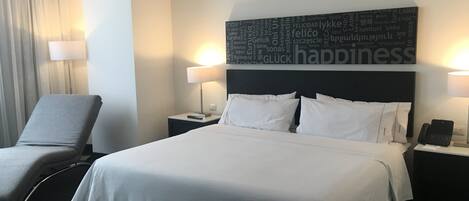 Quarto de hotel com cama, duas mesas de cabeceira, uma cadeira e parede com palavras que expressam felicidade em vários idiomas.